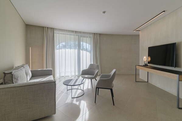 Uma sala de estar moderna com um sofá, duas poltronas, uma mesa de centro, uma TV e uma grande janela com cortinas leves.
