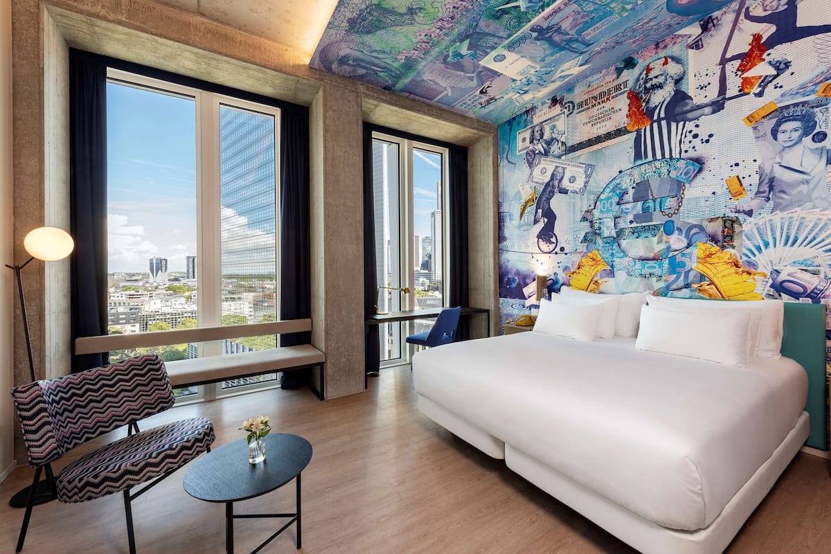 Quarto de hotel moderno com uma cama grande, um banco, uma mesinha com um vaso e vista da cidade através de grandes janelas.