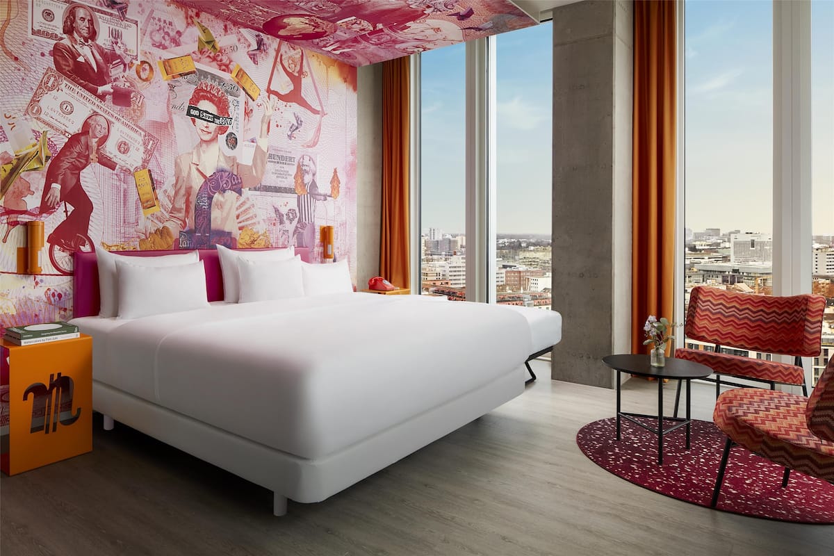 Quarto de hotel moderno com uma cama grande, vista da cidade e um mural colorido na parede.