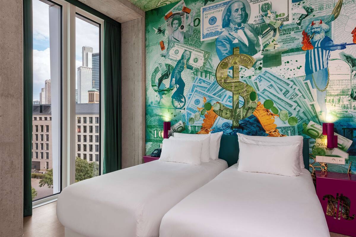 Quarto de hotel com uma grande janela, cama com roupa de cama branca e parede decorada com notas de dinheiro e imagens.