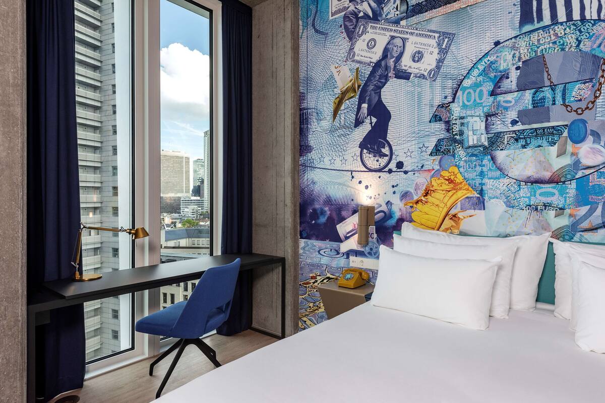 Quarto de hotel com uma grande janela, uma mesa com cadeira, cama com almofadas brancas e um mural colorido na parede.
