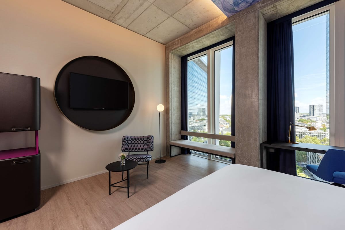 Quarto de hotel com cama, televisão na parede, pequena mesa com cadeira e um banco junto à janela, com vista para a cidade.