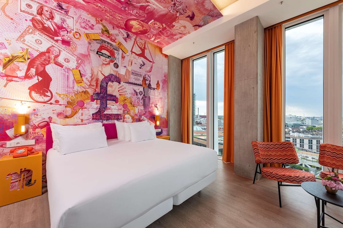Quarto de hotel moderno com uma cama grande, vista para a cidade e um mural vibrante na parede.