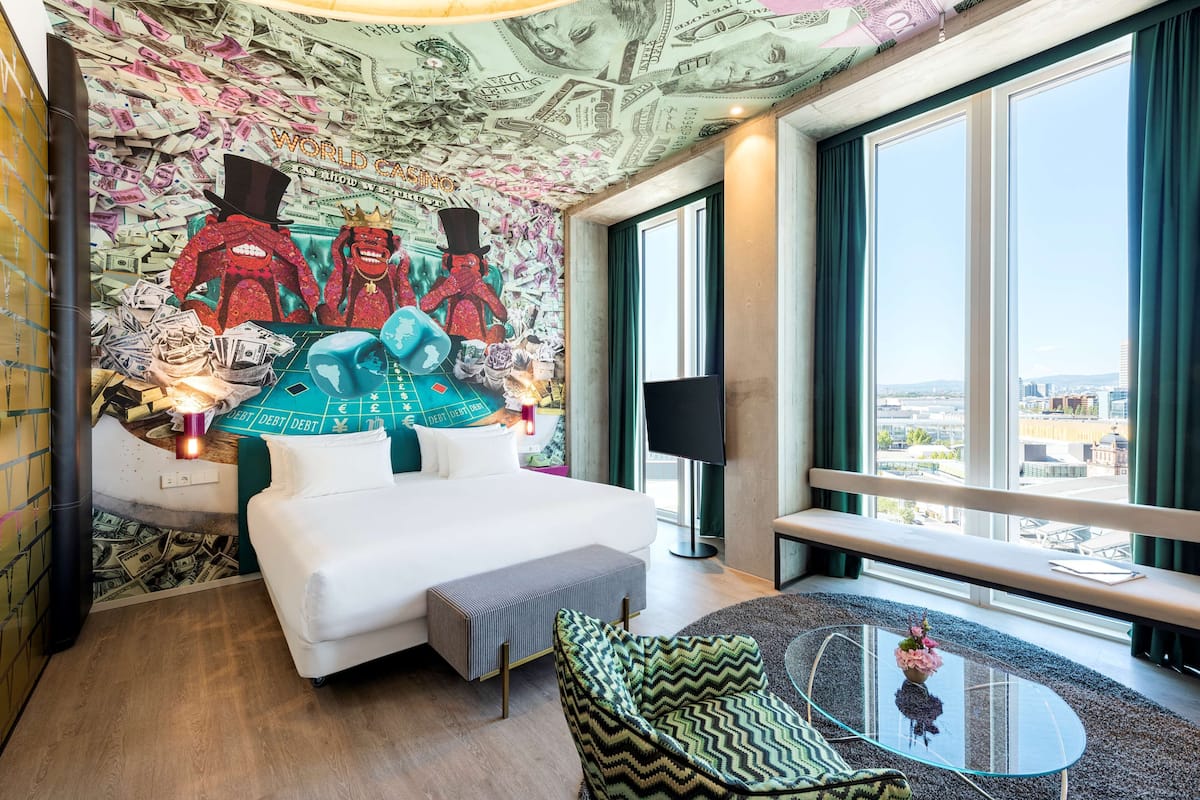 Quarto de hotel com uma cama grande, mesa de centro de vidro, um banco e vista da cidade por grandes janelas.