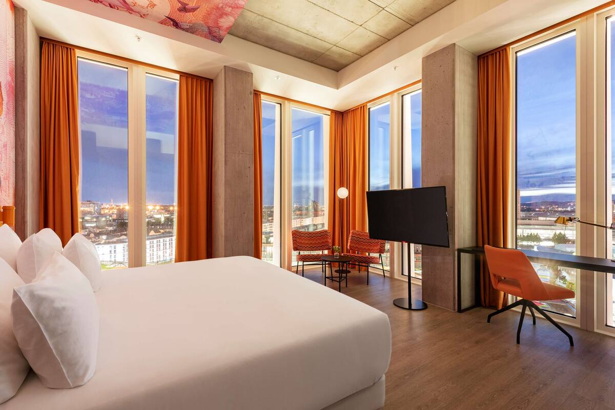 Quarto de hotel com uma cama grande, televisão de tela plana, mesa de jantar com cadeiras e janelas amplas com vista para a cidade.