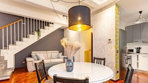 Uma sala de estar moderna com uma mesa de jantar redonda, cadeiras de madeira escura, um sofá bege e um vaso com flores secas. Uma escada com corrimão de madeira leva a um segundo andar.