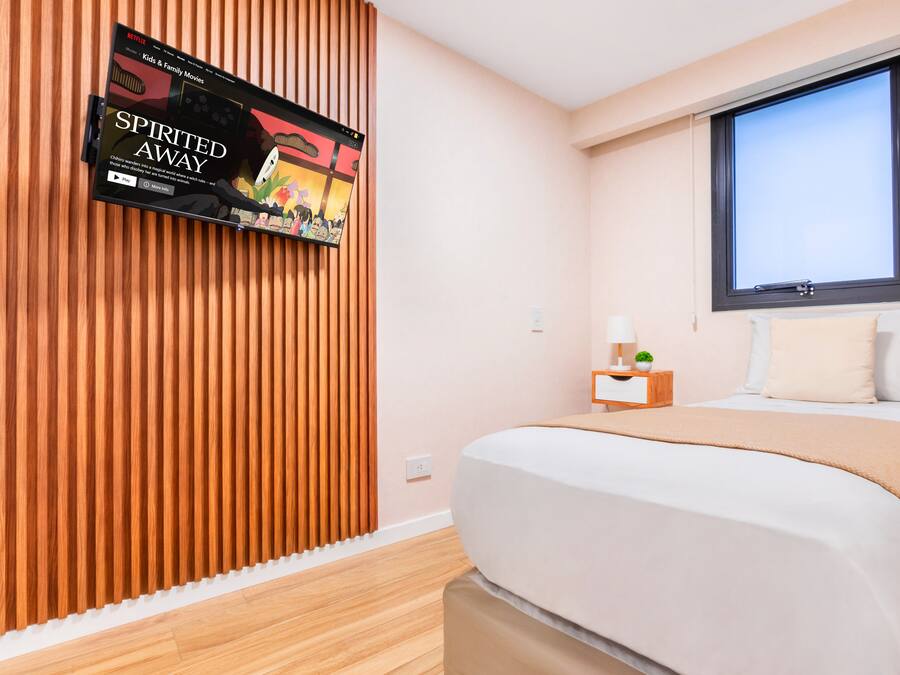 Quarto de hotel com cama, televisão fixada em uma parede revestida de madeira, janela com vista e mesa de cabeceira com abajur.
