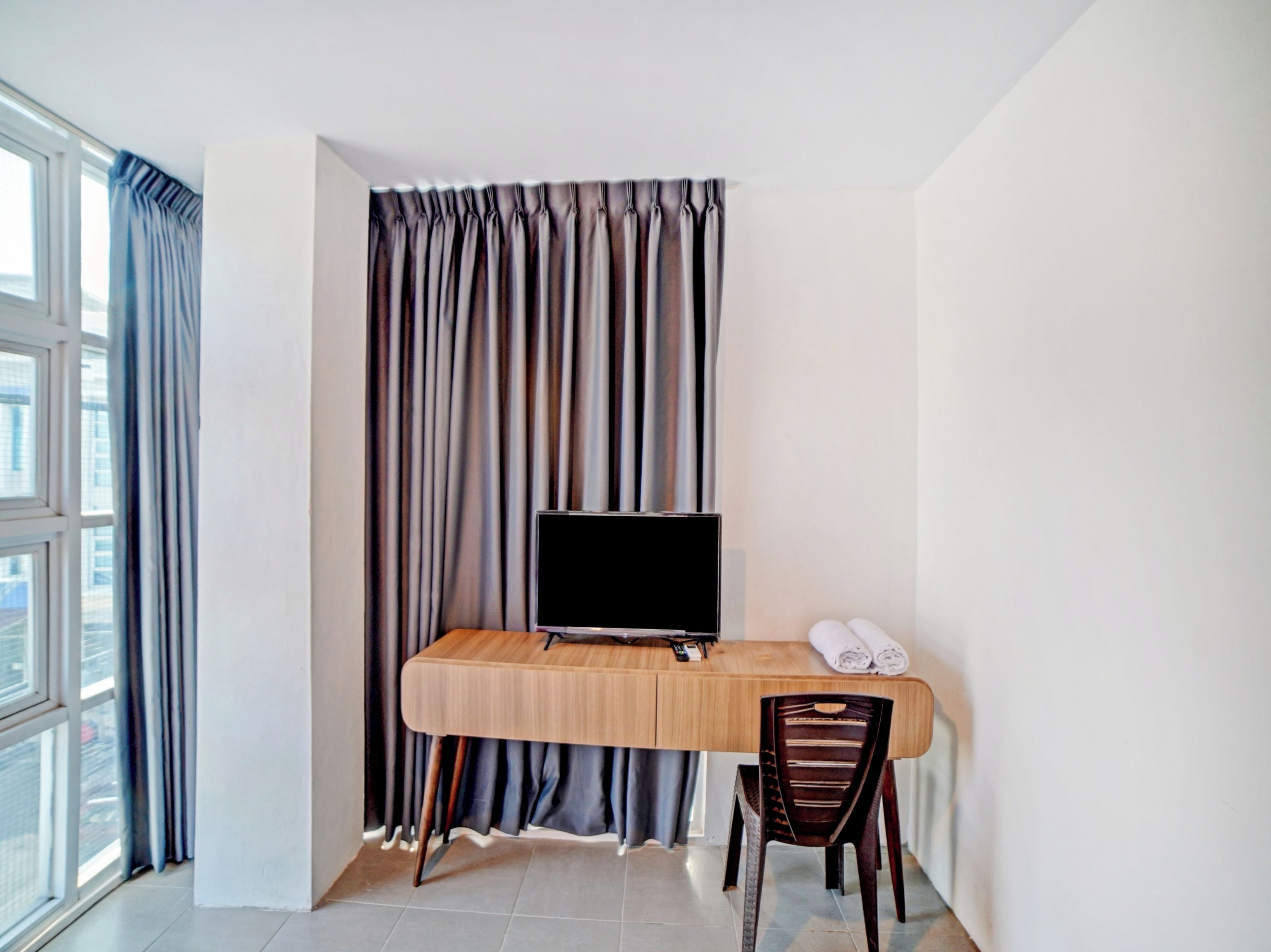 Quarto de hotel com escrivaninha de madeira, televisão, uma cadeira e janela com cortinas.