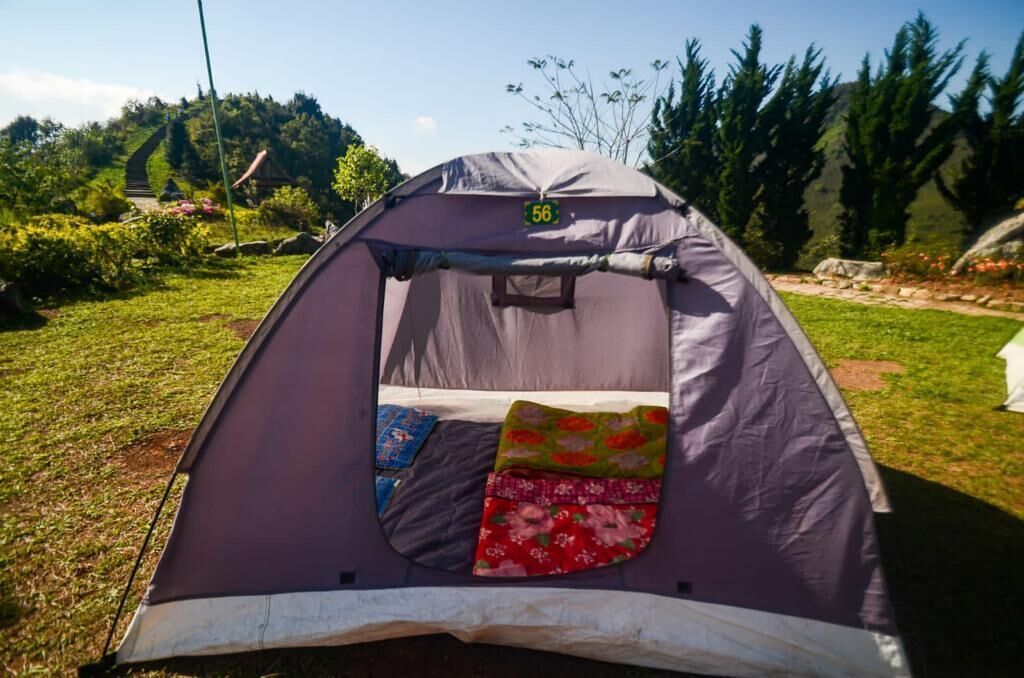 Uma tenda com área de dormir no interior, montada em uma área gramada com árvores e céu claro ao fundo.