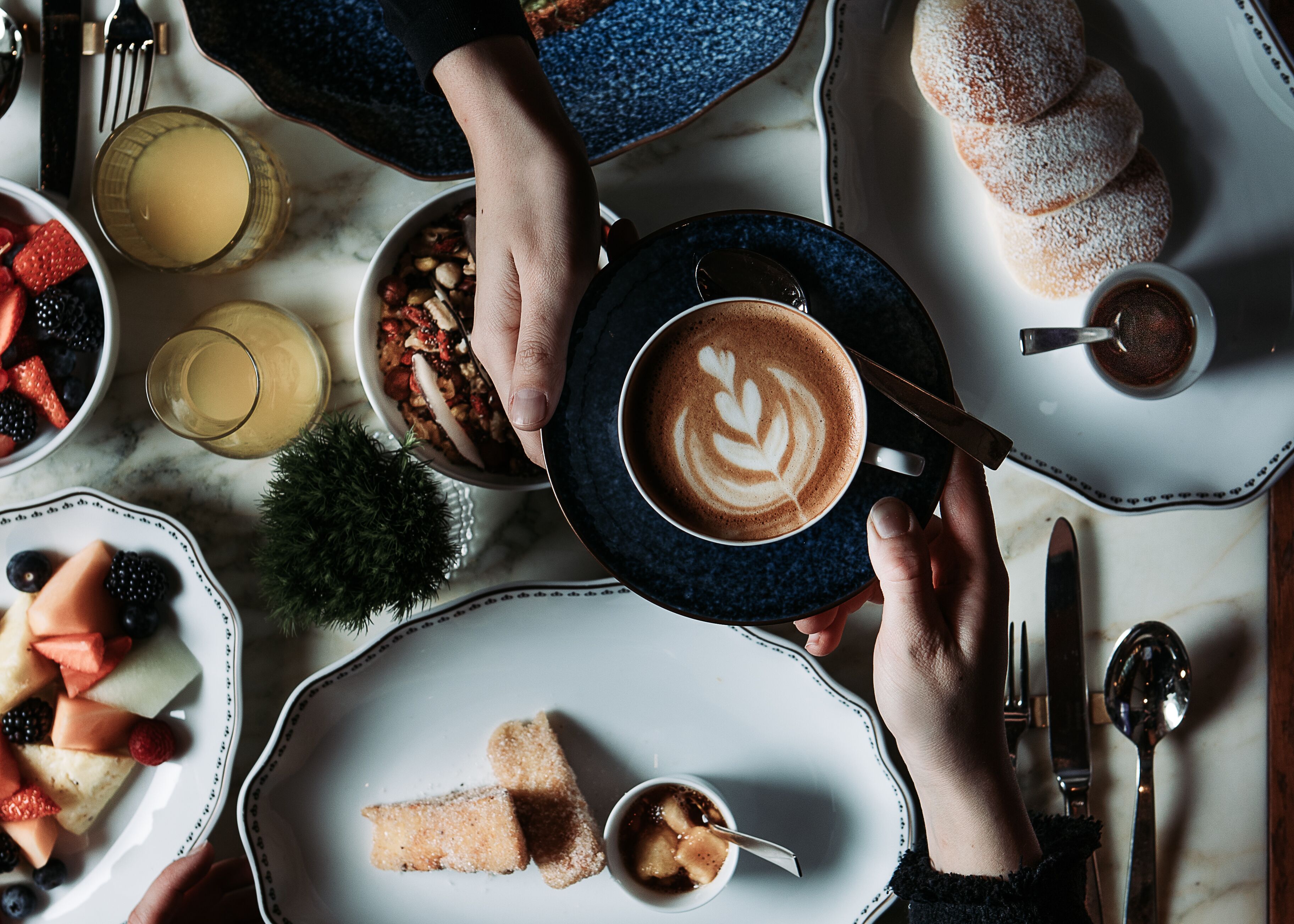 Mesa posta com pratos de pastelaria, uma chávena de café com arte de latte e tigelas de fruta e frutos secos.