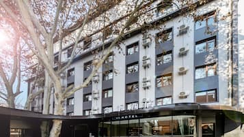 Hotel com fachada moderna, vários aparelhos de ar condicionado e uma grande árvore em frente.