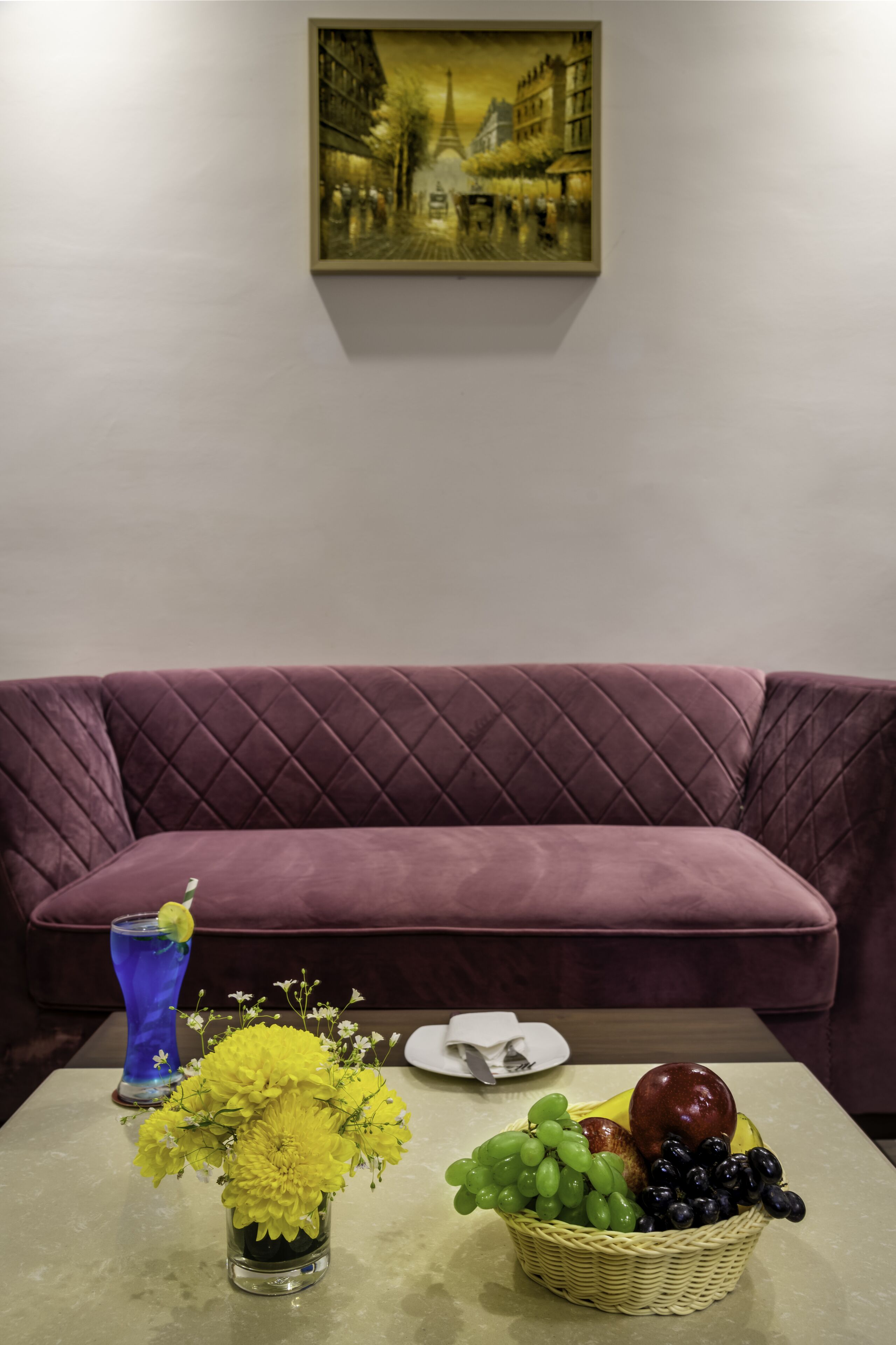 Quarto com área de estar acolchoada, mesa com cesta de frutas e um vaso de flores, e um quadro na parede.