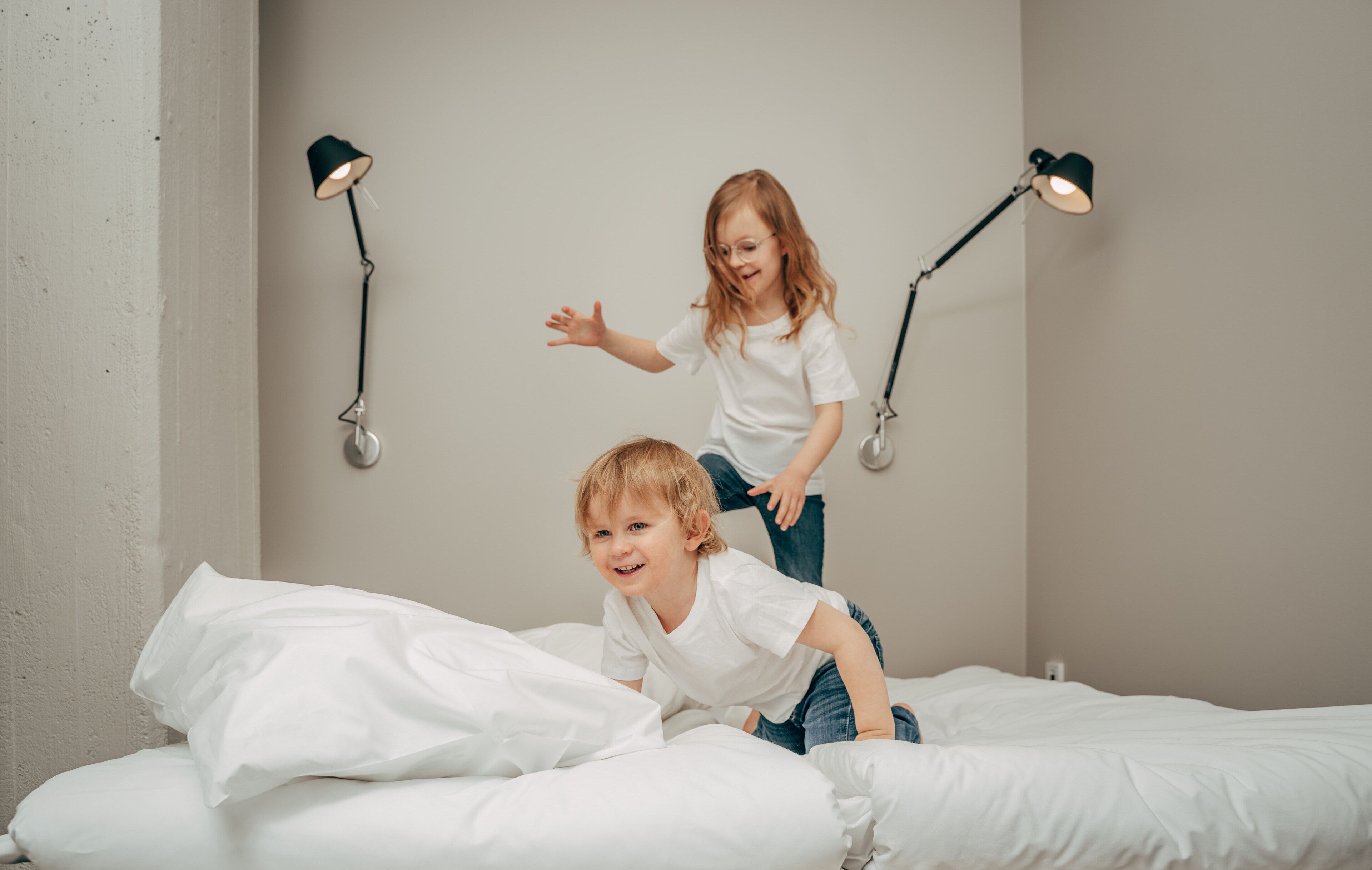 Criança pula em uma cama com roupa de cama branca em quarto com duas luminárias.