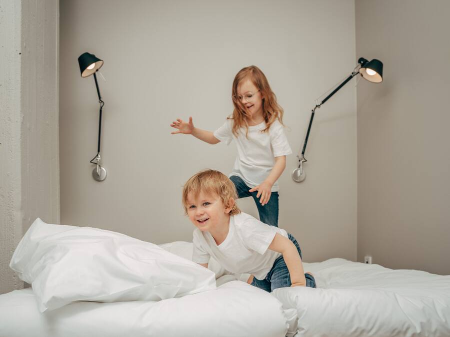 Criança pula em uma cama com roupa de cama branca em quarto com duas luminárias.