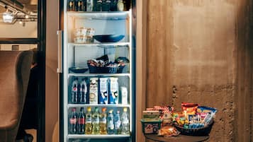 Geladeira compacta com bebidas e lanches, em um quarto moderno com parede de concreto e uma pequena mesa com mais alimentos.