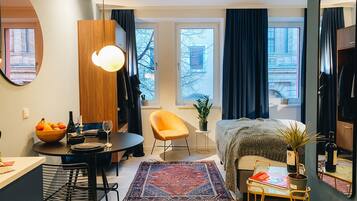 Quarto de hotel moderno com cama, escrivaninha com cadeira, mesa de jantar, espelho grande e vista da cidade pelas janelas.