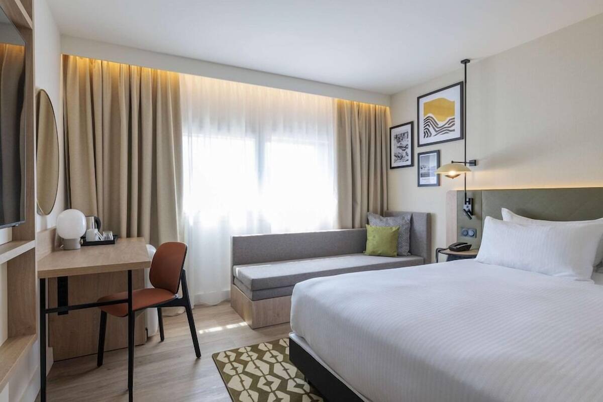Quarto de hotel com cama, escrivaninha, cadeira, sofá e janela com cortinas.