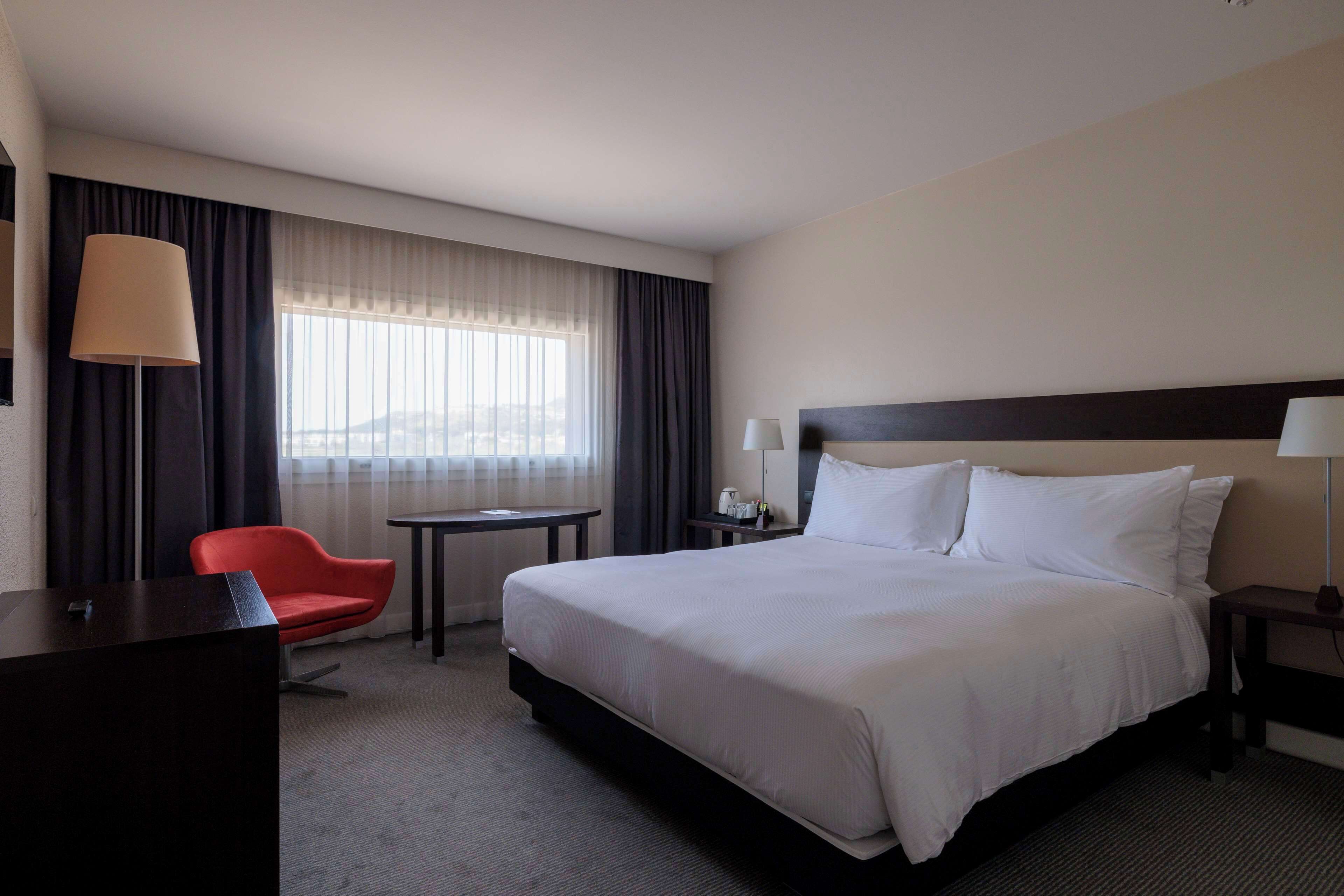Quarto de hotel com uma cama grande, uma escrivaninha, uma cadeira, uma mesinha e uma janela com cortinas.