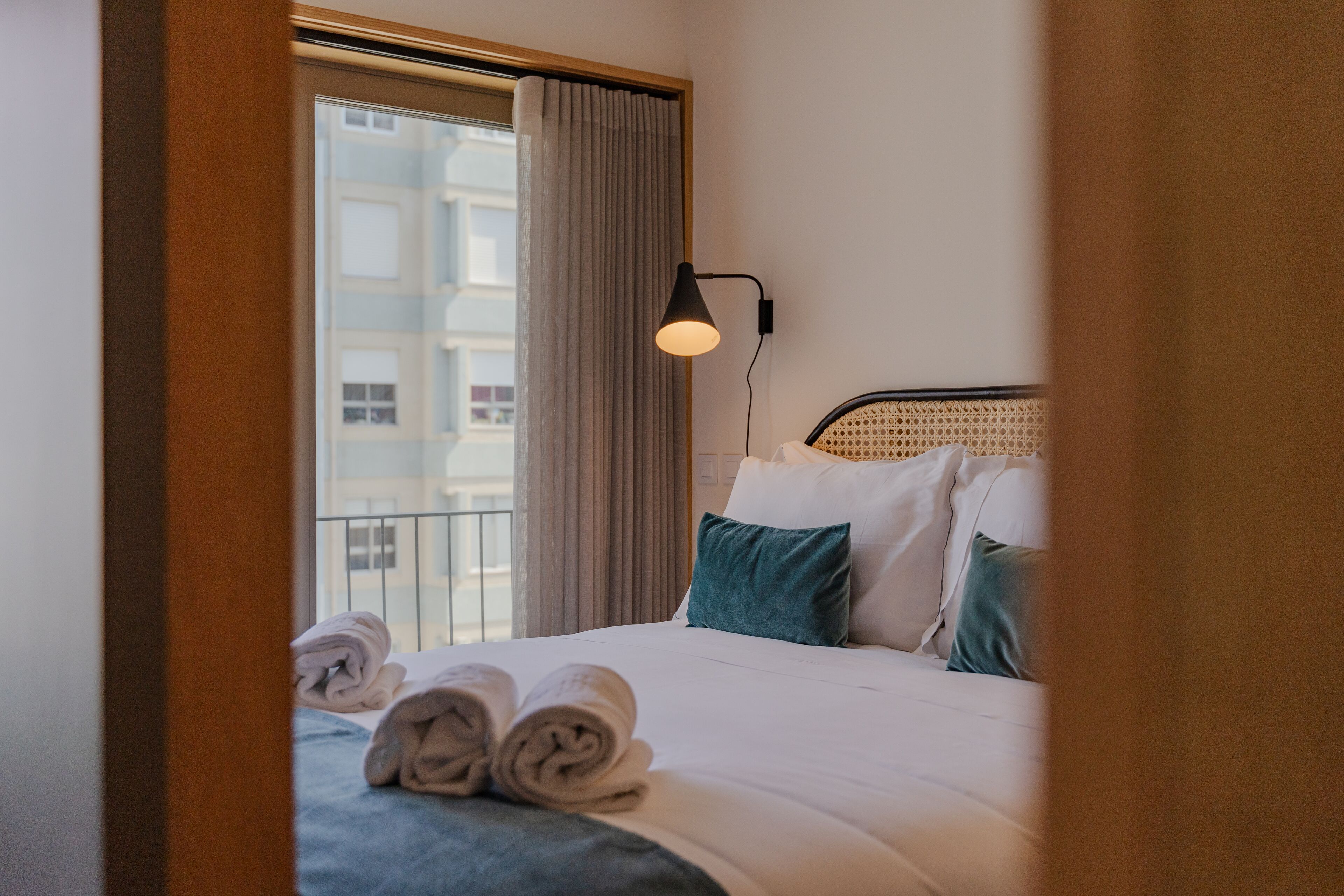 Quarto de hotel com cama, janela com cortinas, luminária fixada na parede e vista para prédios lá fora.
