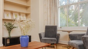 Uma sala de estar com uma mesa de centro de madeira, duas poltronas cinzas, um vaso azul com flores brancas e uma estante com quadros.