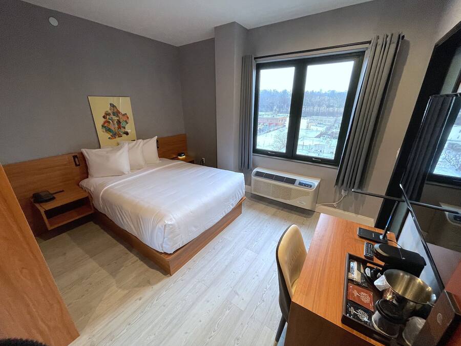 Quarto de hotel com cama, secretária com máquina de café, janela com vista e aquecedor fixado na parede.