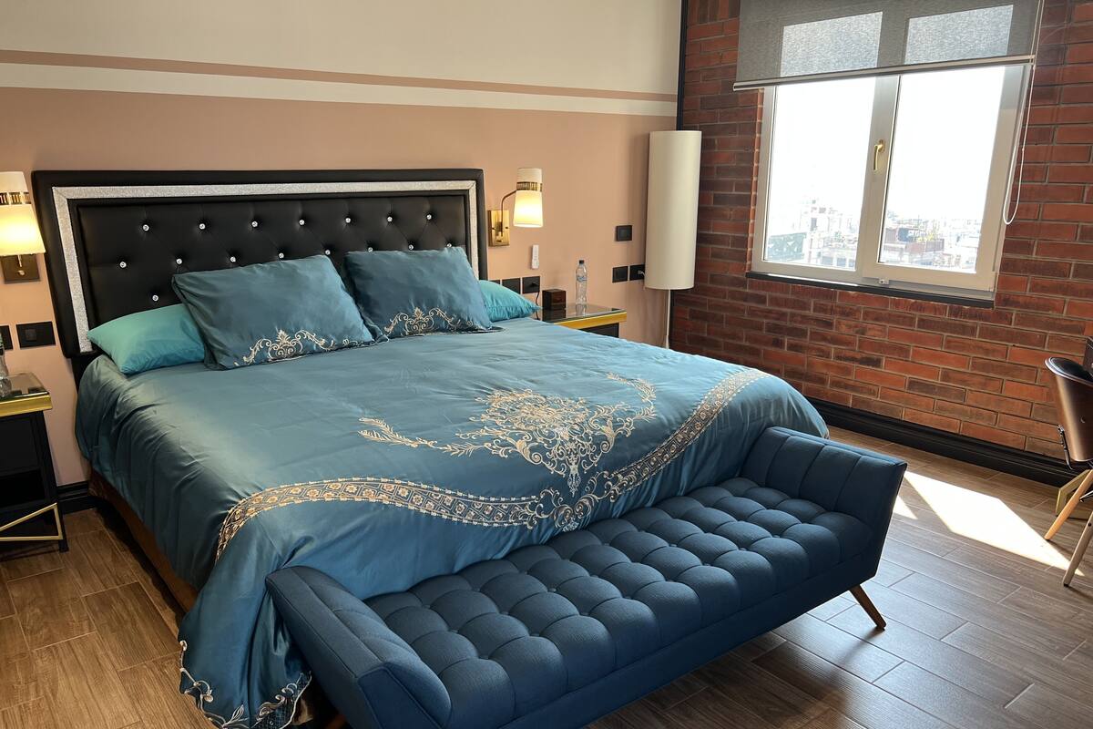 Quarto moderno com uma cama grande, um banco e vista da cidade pela janela.