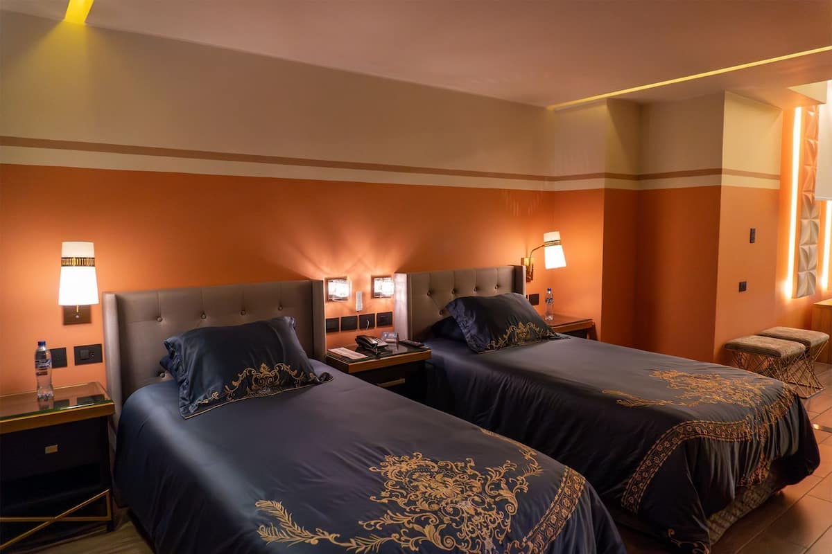Quarto de hotel com duas camas, cada uma com cabeceira, mesas de cabeceira, luminárias e um pequeno banquinho.