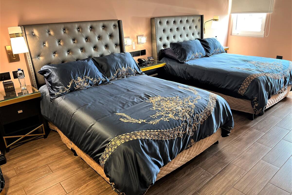 Quarto de hotel com duas camas, cada uma com cabeceira estofada, roupa de cama azul-escuro com bordado dourado e piso de madeira.