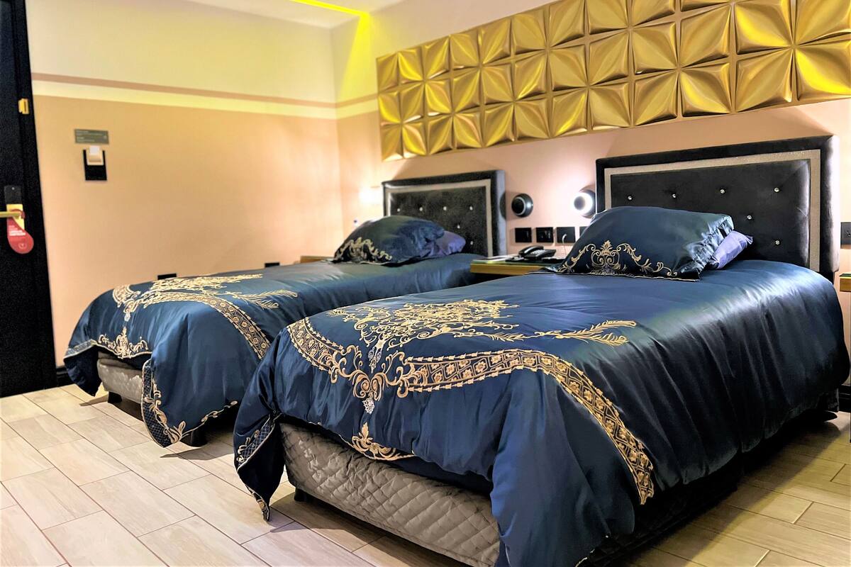 Quarto de hotel com duas camas, cada uma com colcha azul escura e bordado dourado, cabeceira moderna com padrão geométrico e luminária no teto.