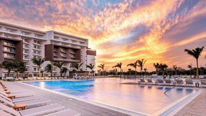 Área de piscina de hotel com céu ao pôr do sol, palmeiras e espreguiçadeiras.