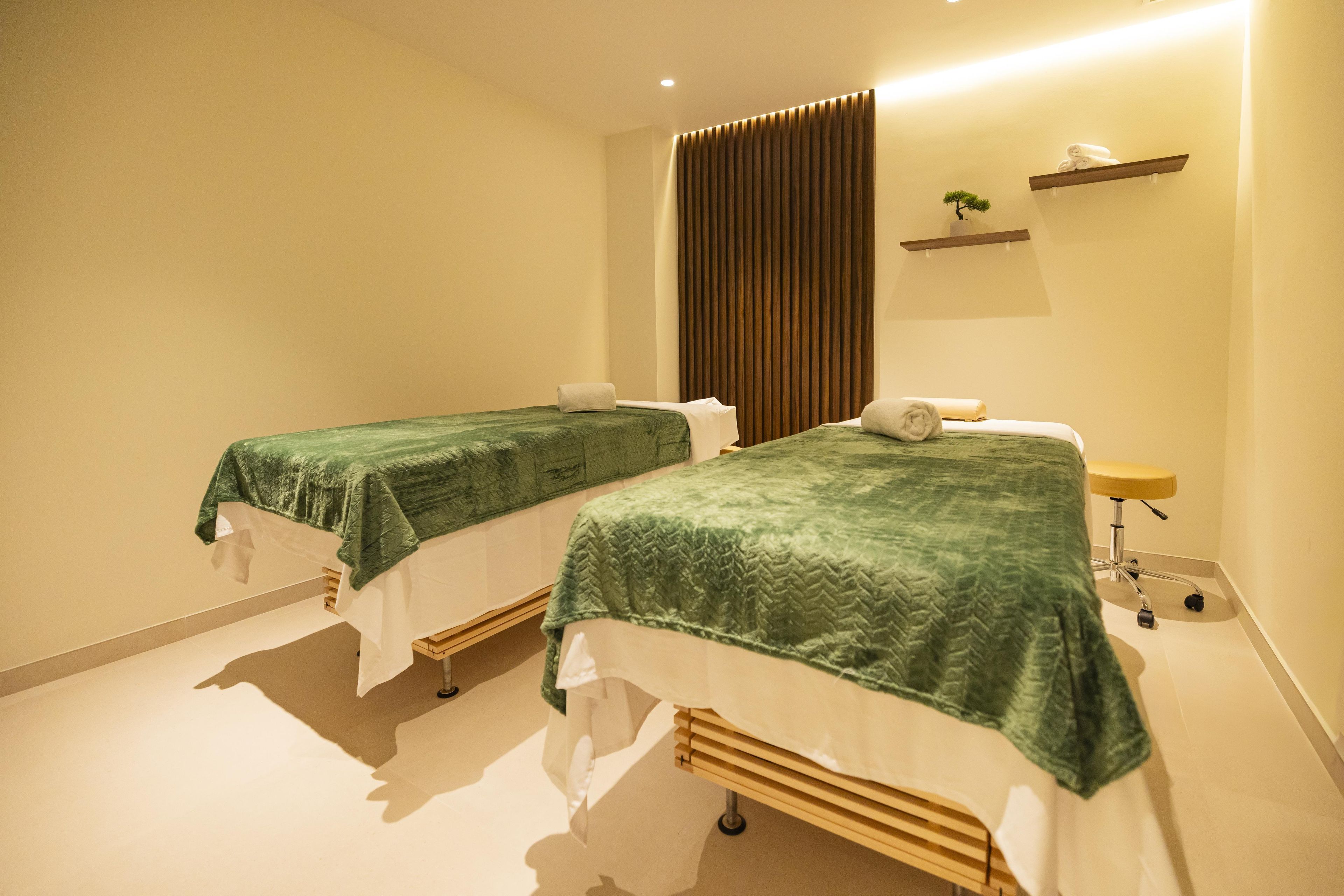 Quarto com duas camas, cada uma com um cobertor verde, uma pequena mesinha de cabeceira e uma prateleira na parede com toalhas.