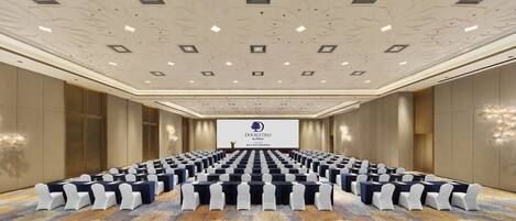 Uma sala de conferências com fileiras de mesas e cadeiras, uma grande tela exibindo o logotipo da DoubleTree by Hilton e um tapete estampado.