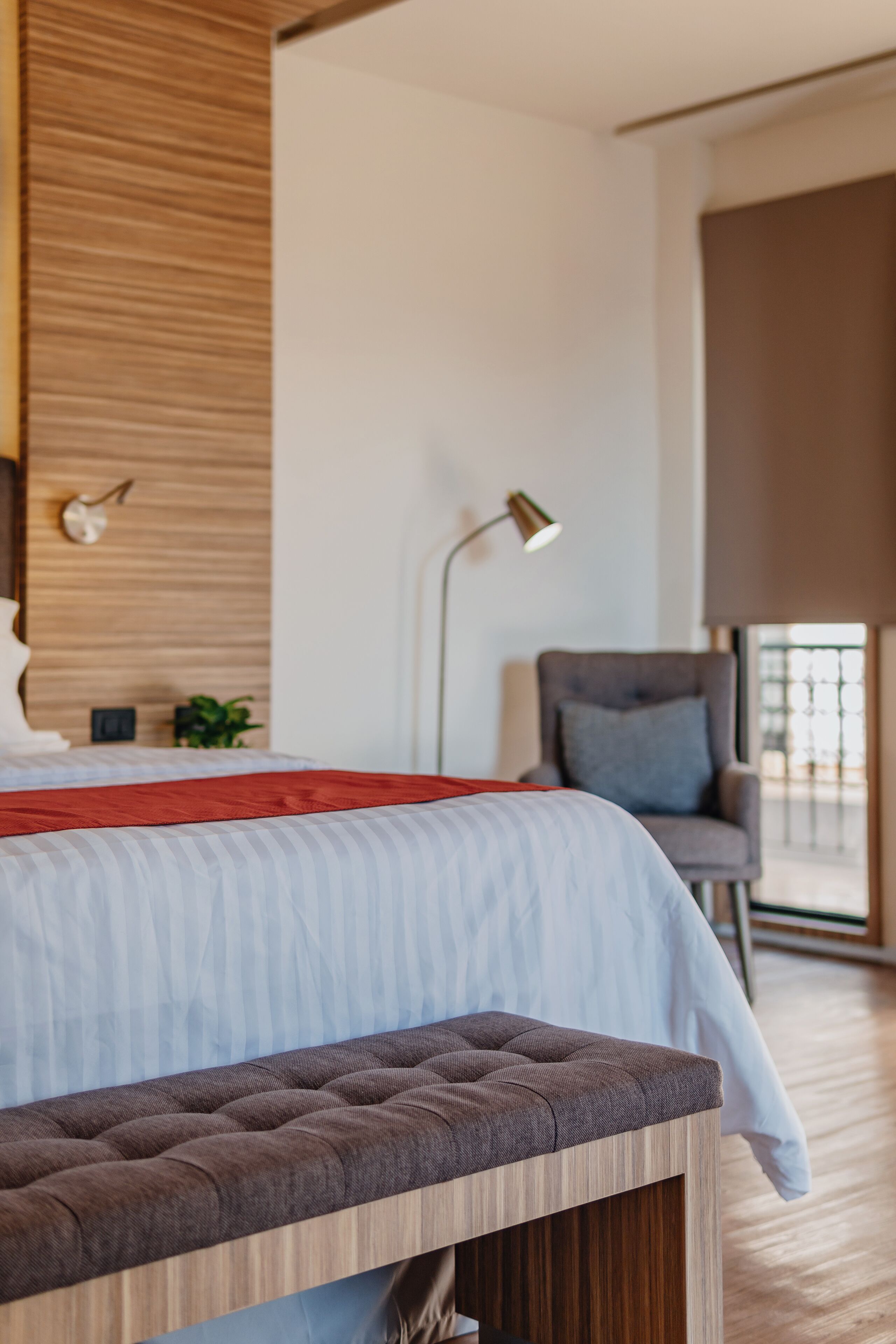 Quarto moderno com cama, um banquinho, uma cadeira e uma mesinha de cabeceira.