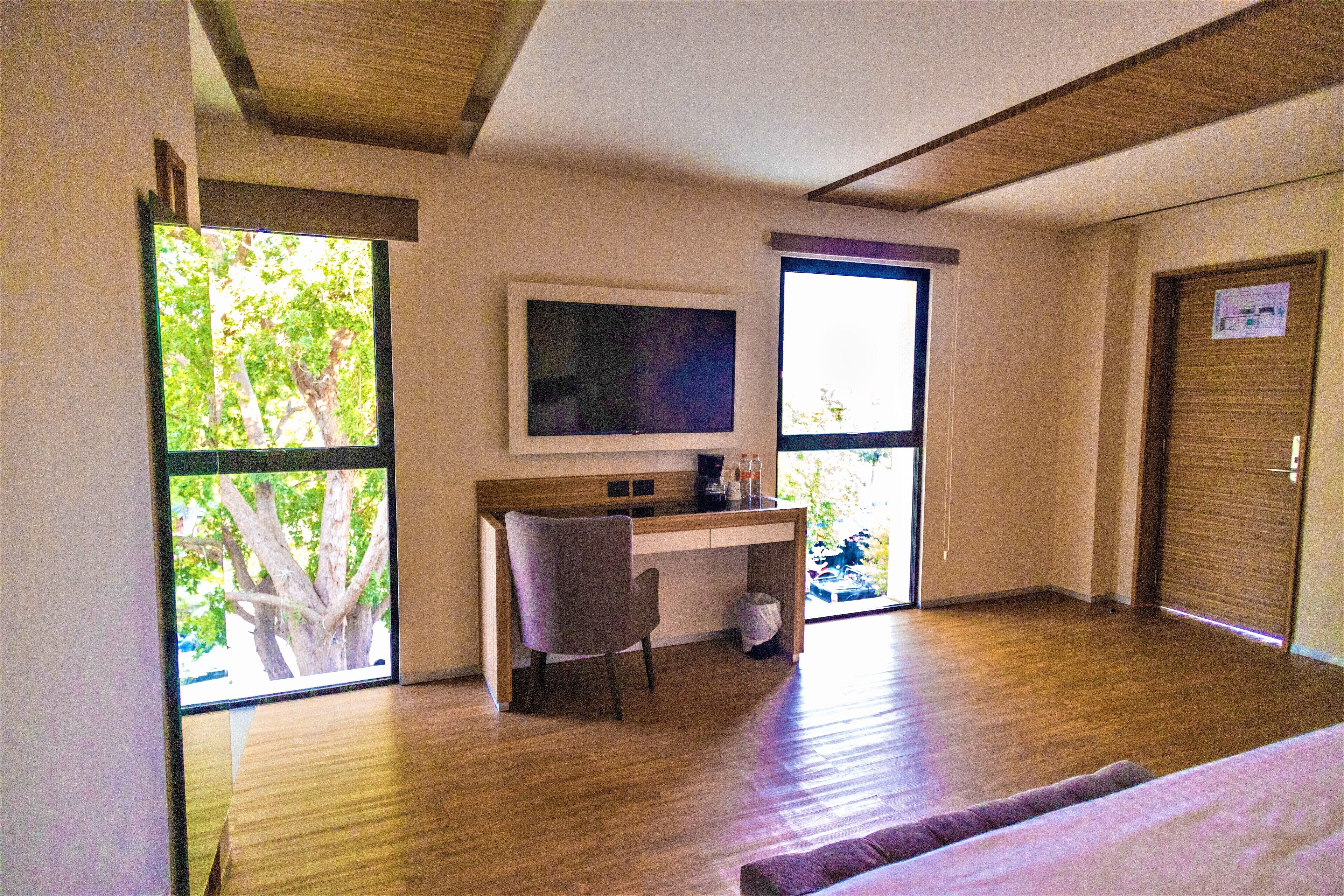 Quarto com escrivaninha de madeira, cadeira e televisão. Há uma janela grande com vista para árvores e uma porta com tranca.