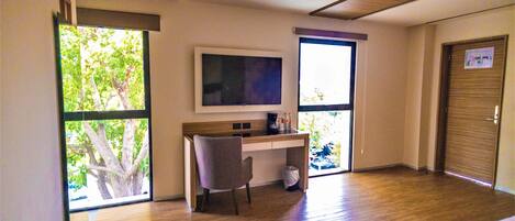 Quarto com escrivaninha de madeira, cadeira e televisão. Há uma janela grande com vista para árvores e uma porta com tranca.