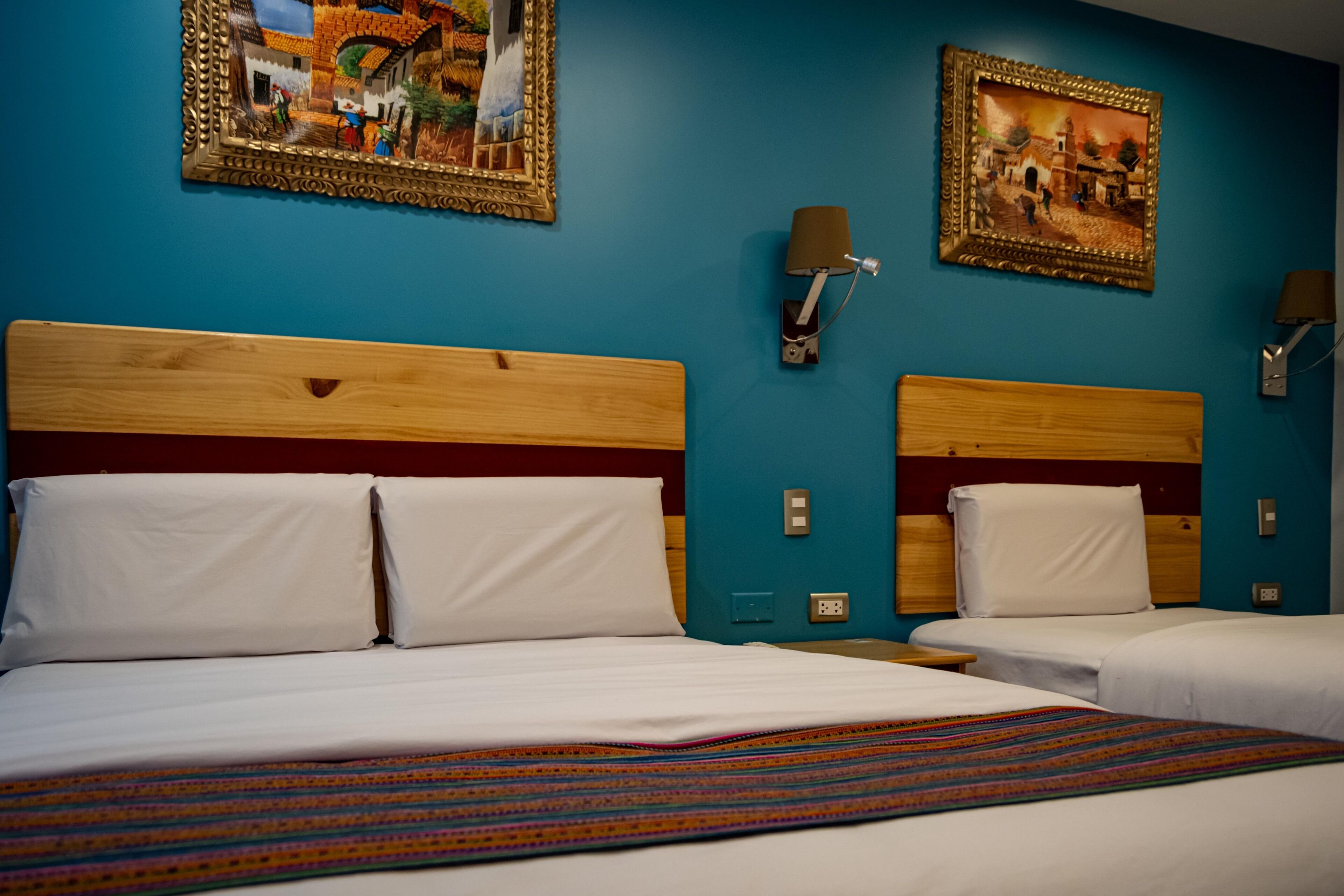 Quarto de hotel com duas camas, cabeceiras de madeira, roupa de cama branca e duas imagens emolduradas na parede.
