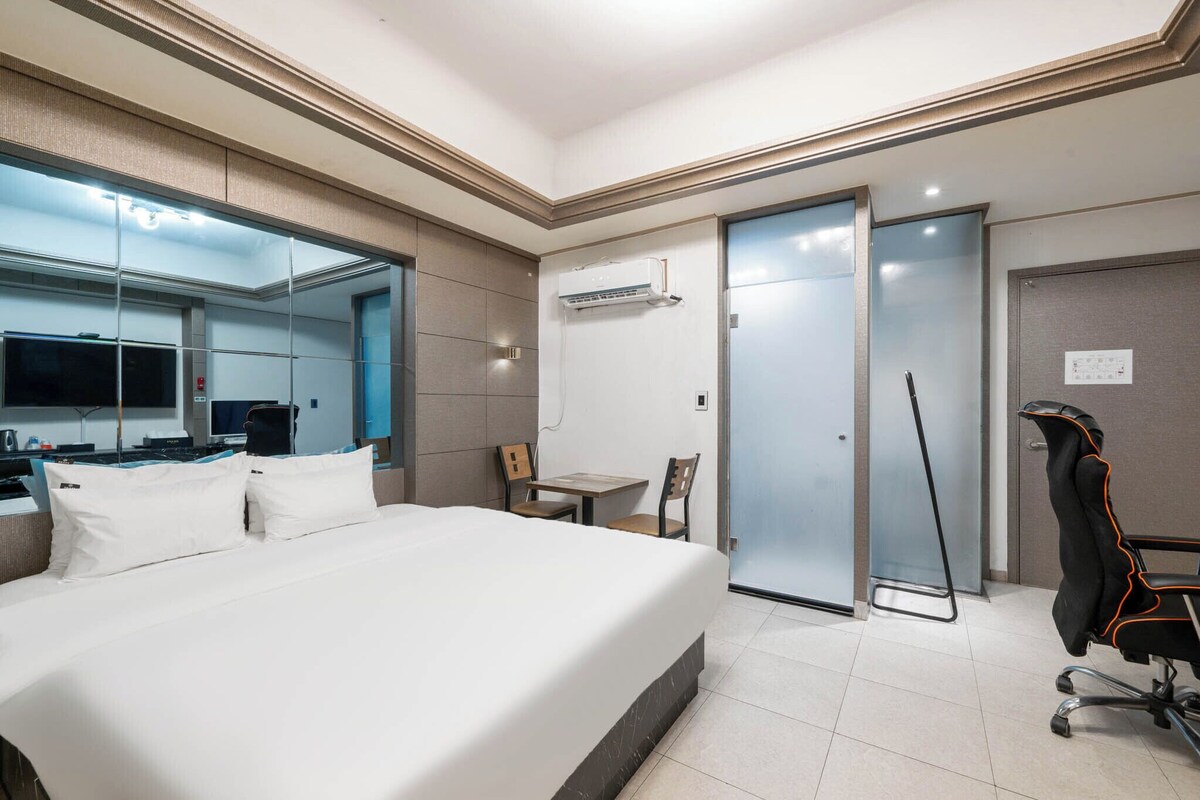 Quarto de hotel com cama, escrivaninha, cadeira, espelho e porta.