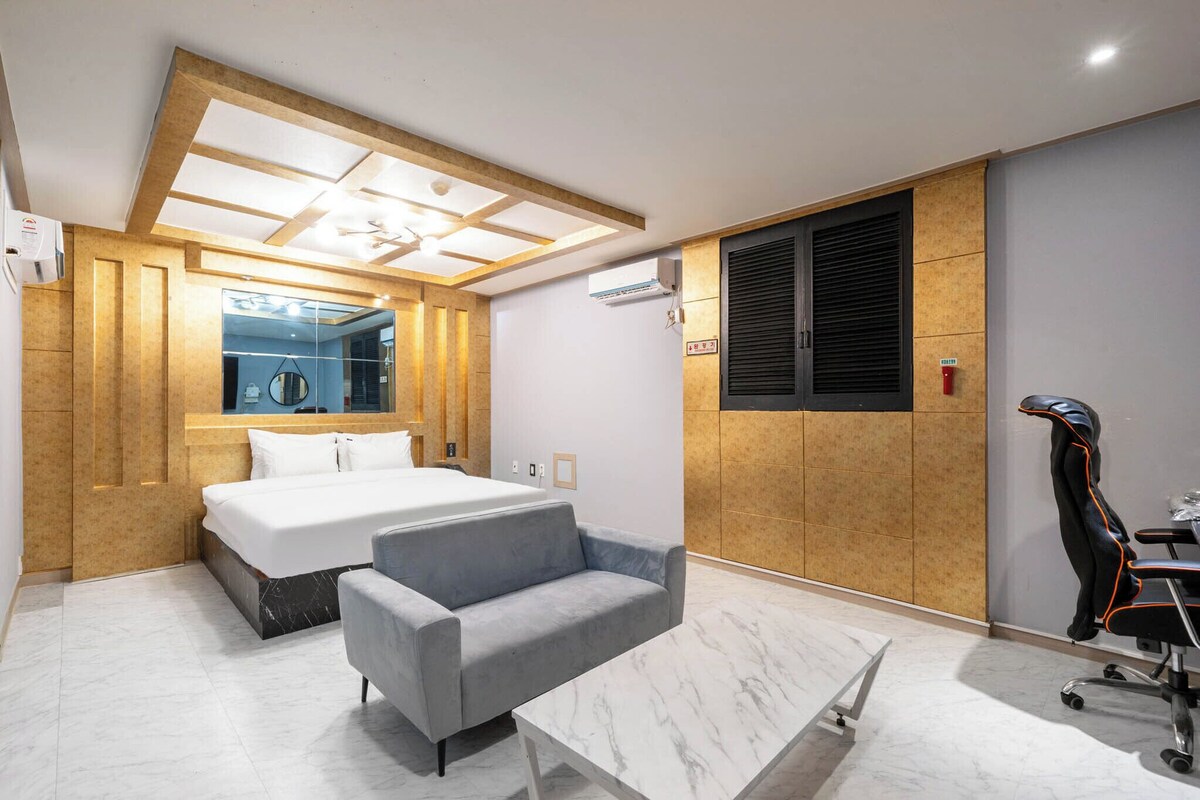 Quarto de hotel moderno com cama, sofá cinza, mesa de centro de mármore, cadeira de escritório preta, ventoinha de teto e um espelho grande.