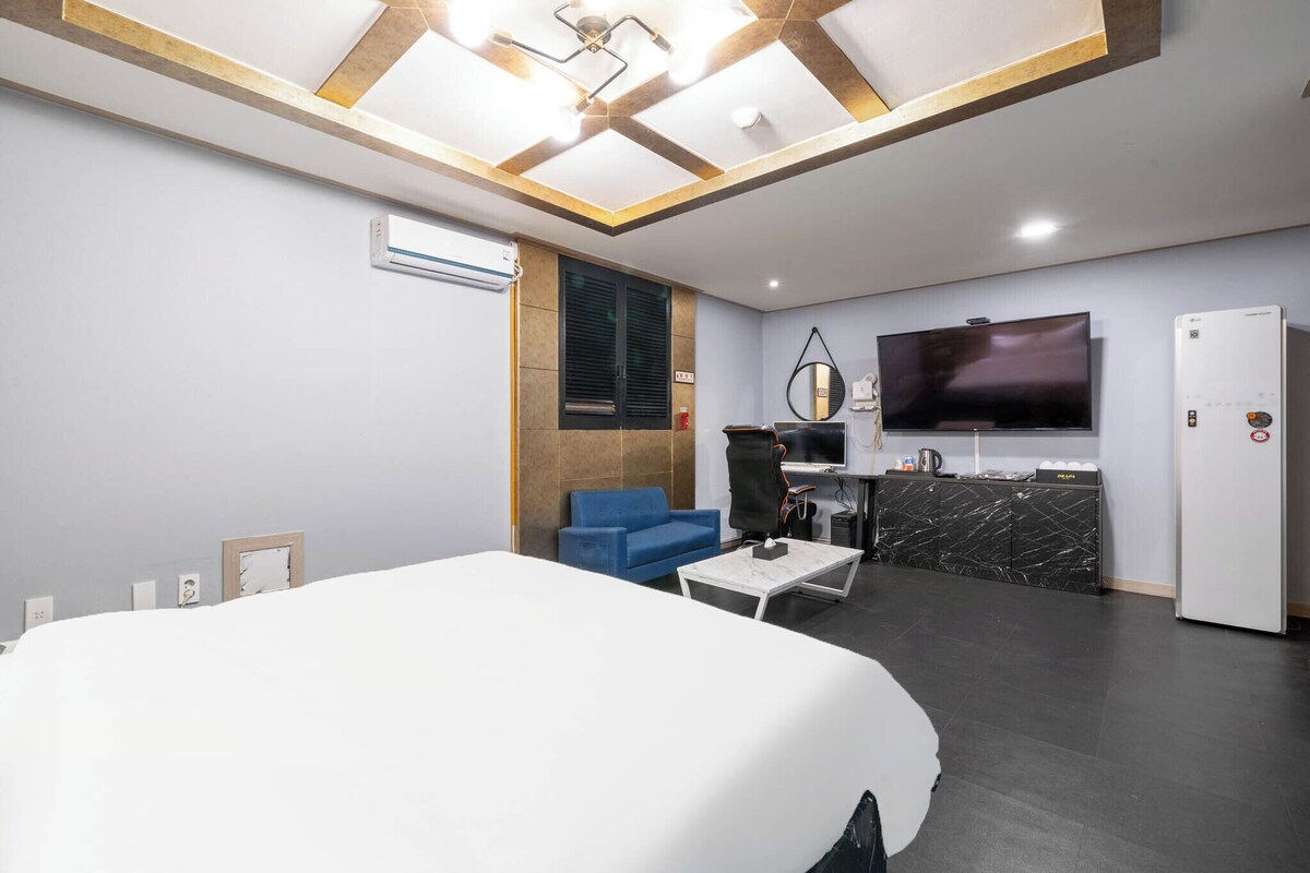 Quarto de hotel com cama, uma cadeira azul, mesa de centro, TV, frigorífico e área de cozinha com bancada e pia.
