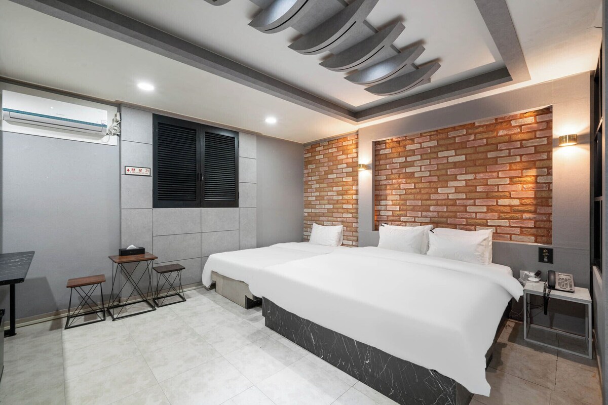 Quarto de hotel moderno com uma cama grande, parede decorada com tijolos, uma pequena escrivaninha e uma cadeira.