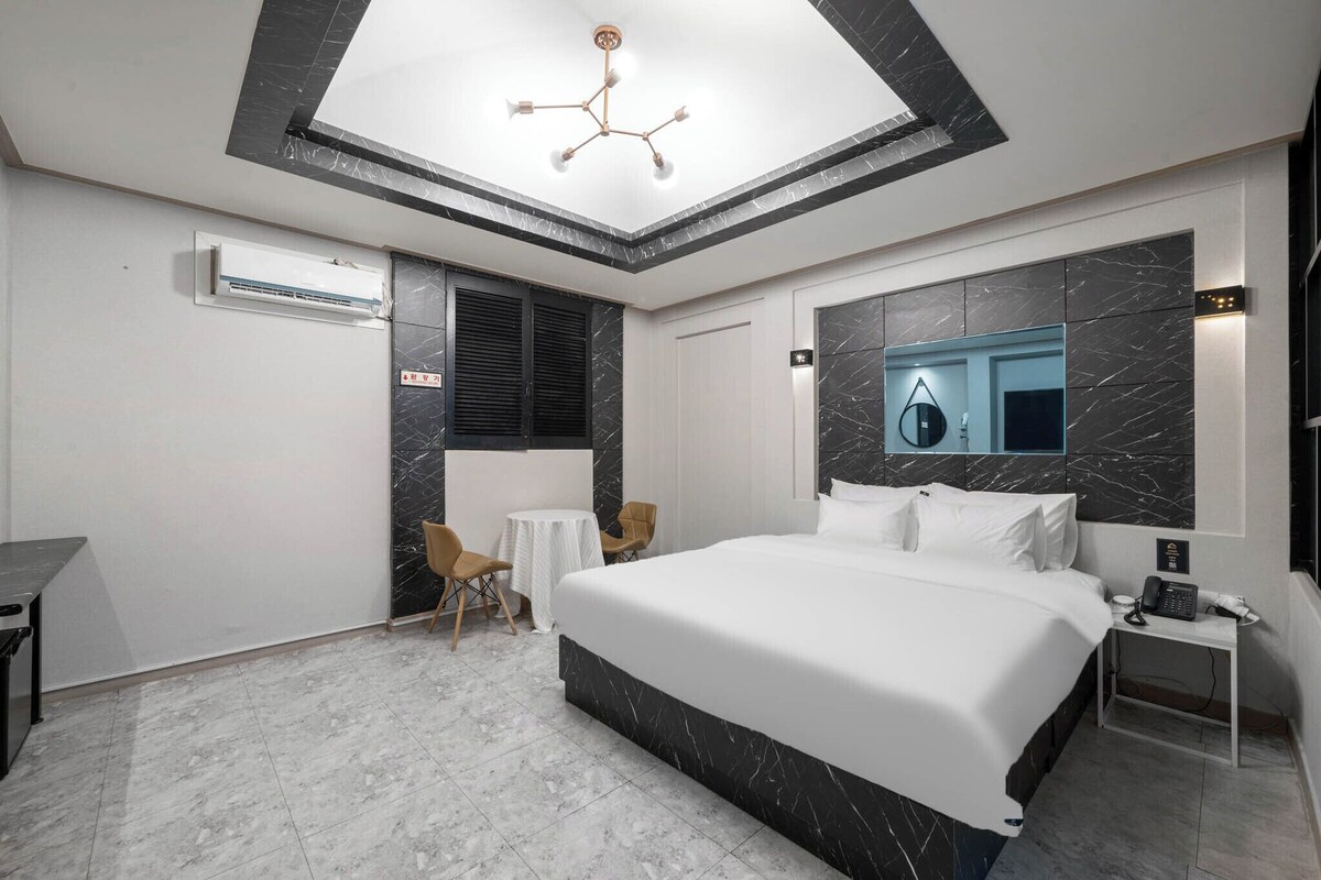 Quarto de hotel moderno com uma cama grande, uma escrivaninha, uma cadeira, uma mesa pequena e um lustre.