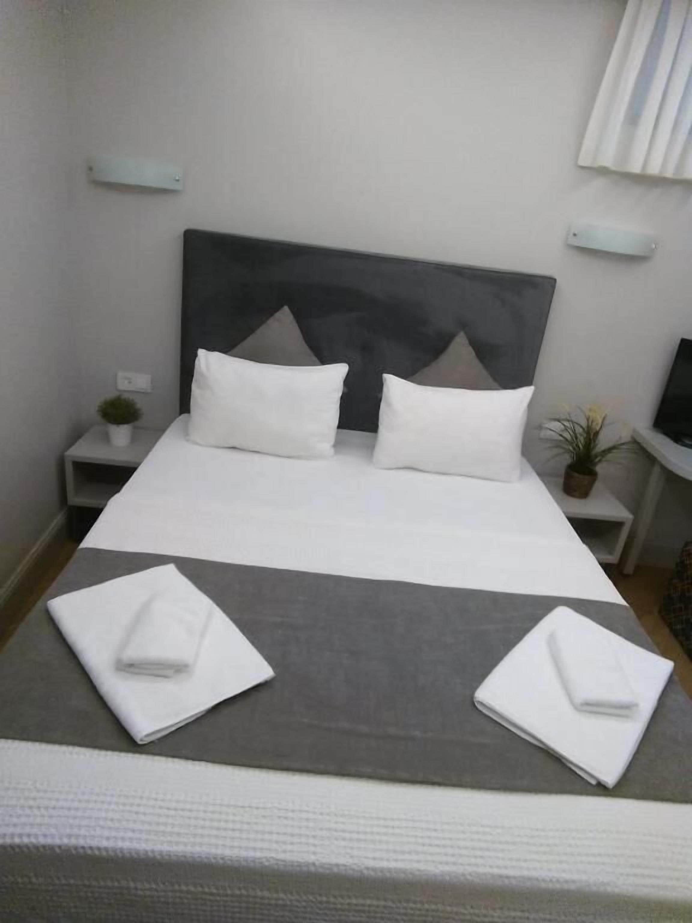 Cama bem arrumada com cabeceira cinza, dois travesseiros brancos e toalhas brancas dobradas sobre a cama.