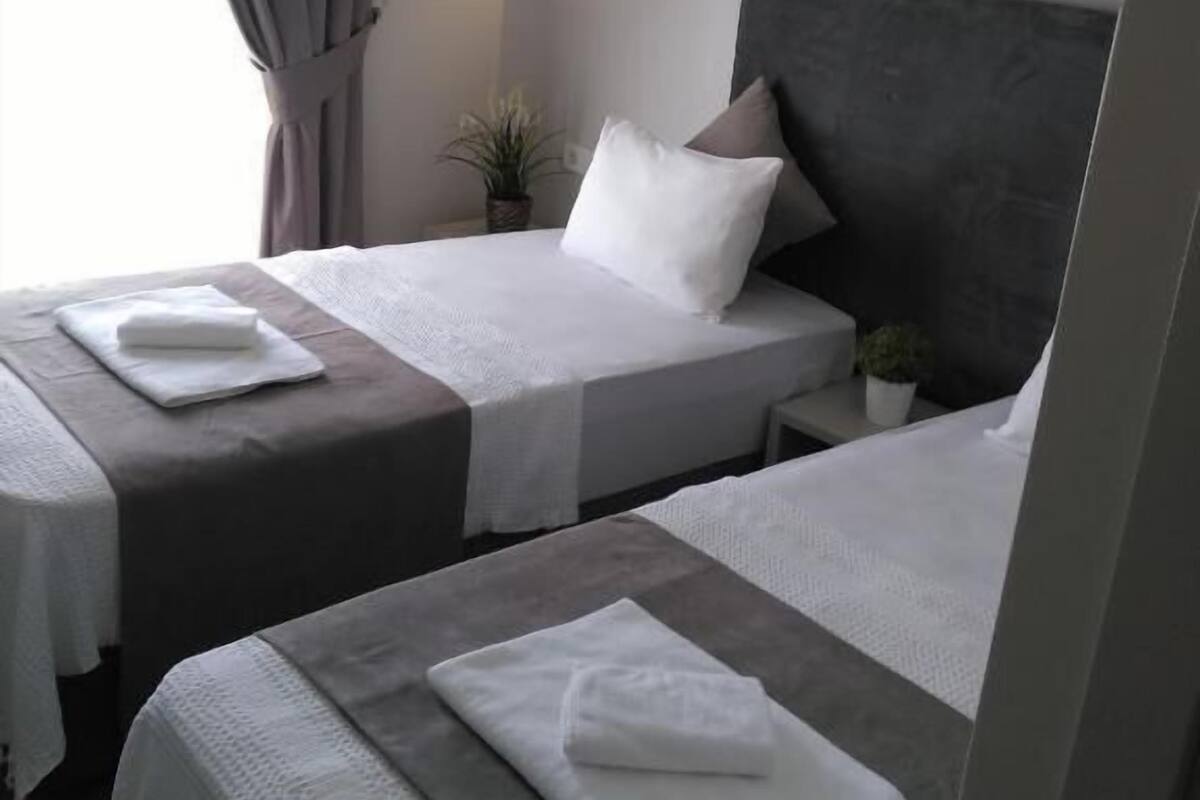 Quarto de hotel com duas camas, ar-condicionado fixado na parede, uma pequena planta em vaso e uma janela com cortinas.