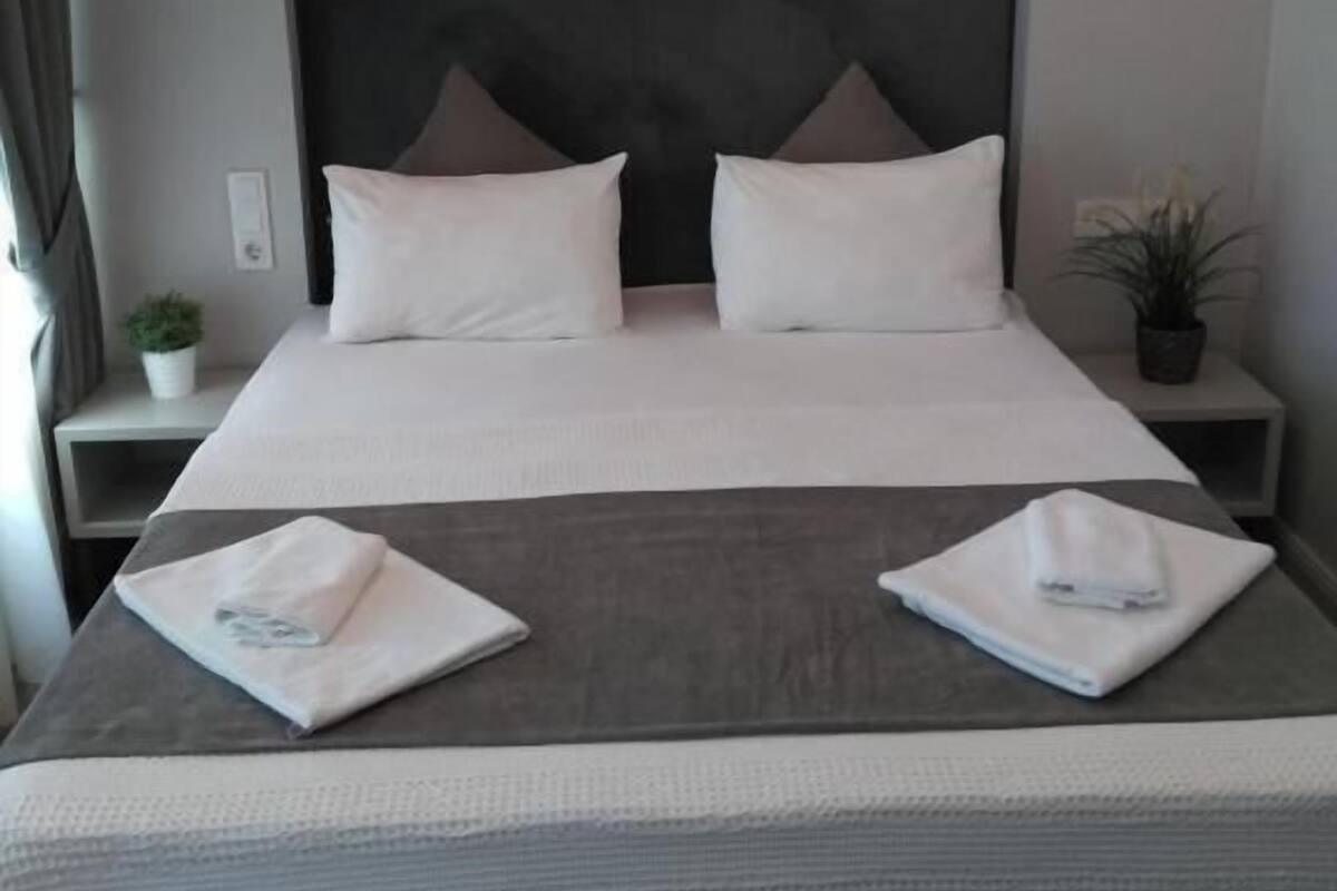Cama bem arrumada com cabeceira escura, dois travesseiros e duas toalhas dobradas.
