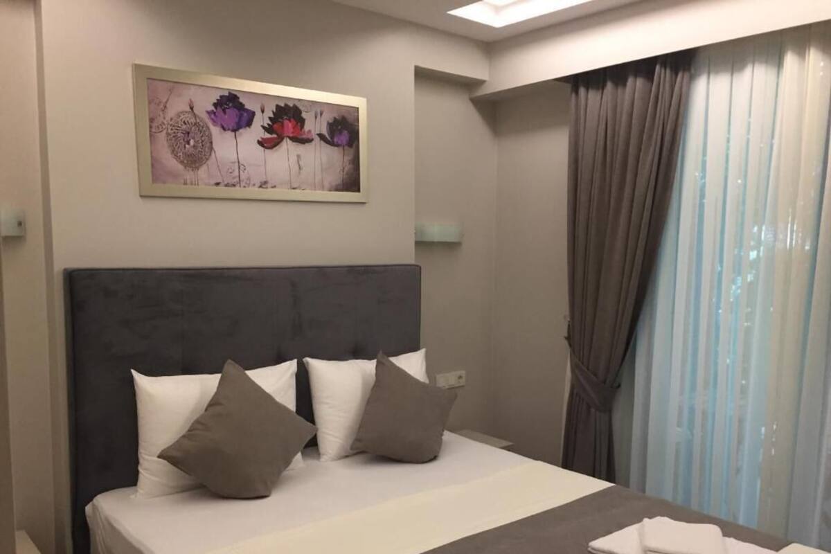 Uma cama bem arrumada com roupa de cama branca e castanha, uma obra de arte floral emoldurada acima dela, e uma janela com cortinas.