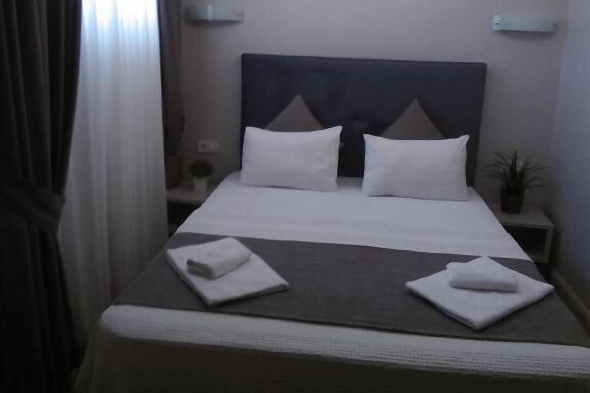 Quarto de hotel com cama, duas mesas de cabeceira, um criado-mudo, janela com cortinas e um claraboia.