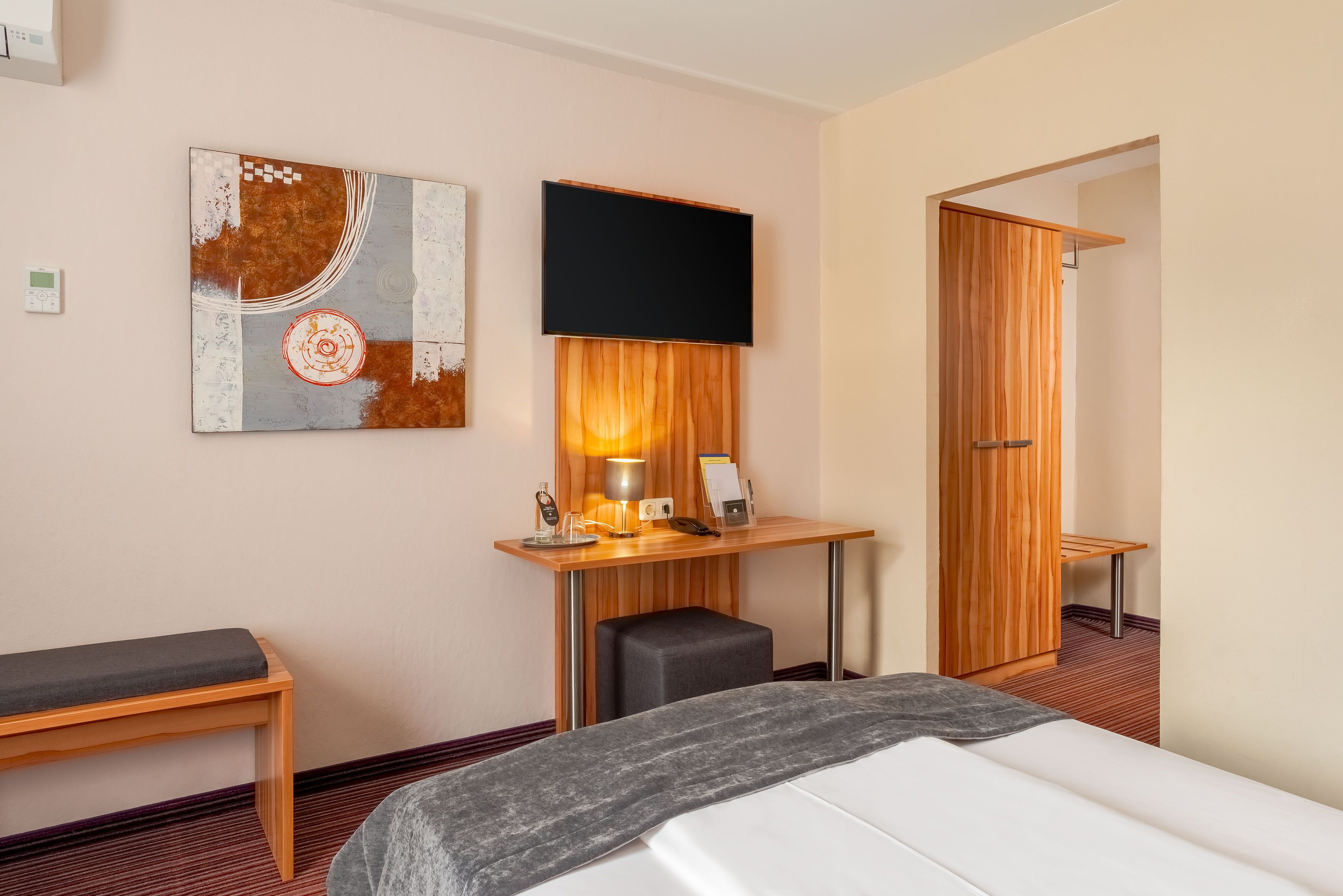 Quarto de hotel com uma cama, uma mesa com televisão de tela plana, um banco e uma obra de arte na parede.