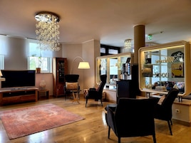 Uma sala de estar moderna com televisão de tela plana, um armário de madeira, uma poltrona preta, uma vitrine de vidro com objetos decorativos e um grande lustre.