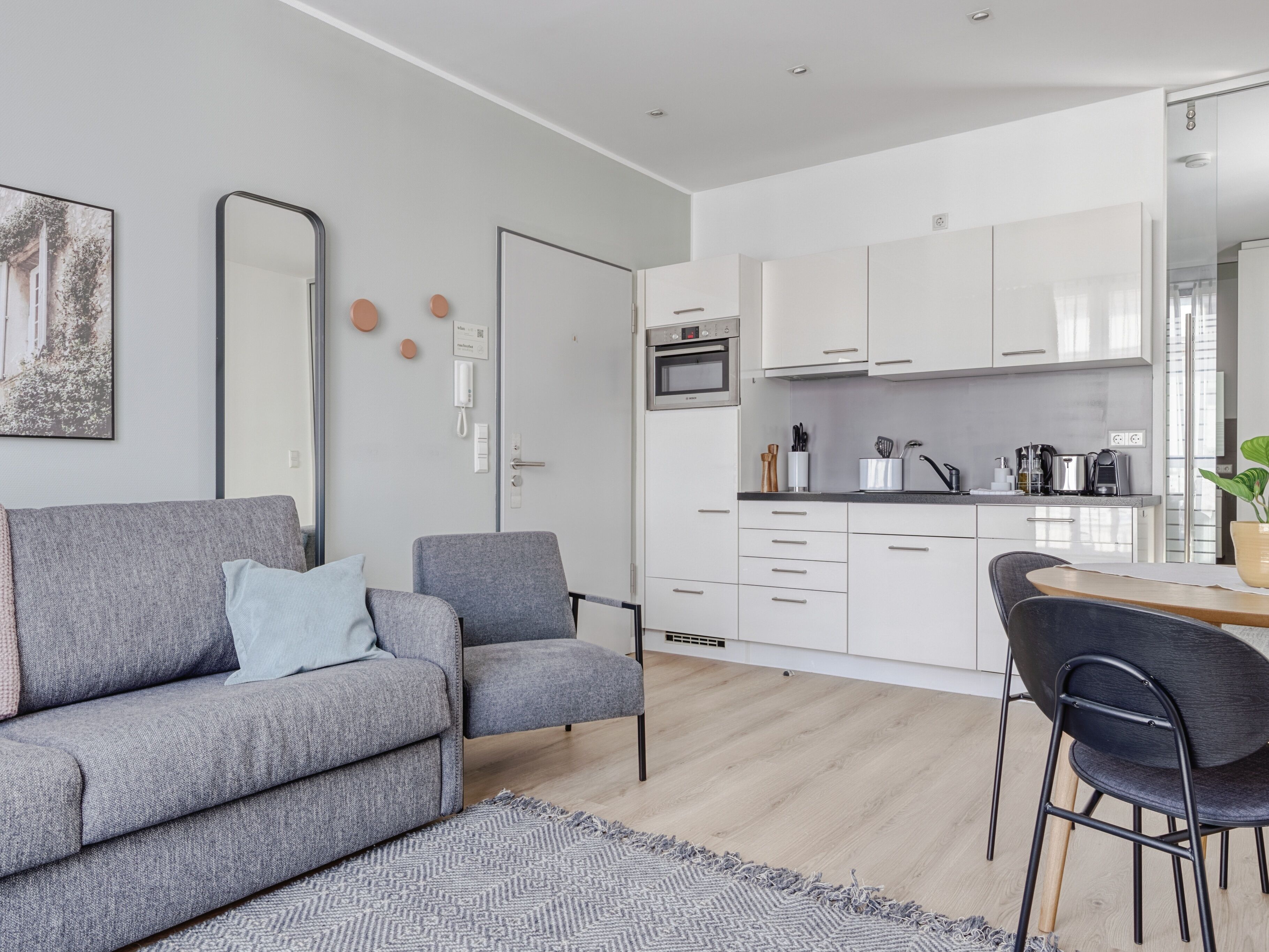 Uma sala de estar moderna com um sofá cinzento, uma poltrona pequena e uma área de jantar com mesa e cadeiras de madeira.