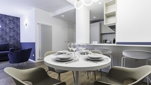 Uma sala de jantar moderna com uma mesa redonda posta para quatro pessoas, rodeada por quatro cadeiras. Uma área de cozinha com bancada branca e chaleira é visível ao fundo.